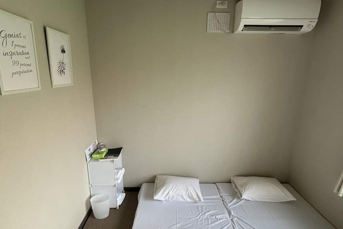Quarto de hotel com uma cama, uma pequena escrivaninha, ar condicionado e duas fotografias emolduradas na parede.