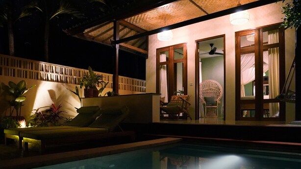 Vista noturna de uma área de piscina com espaço de estar coberto, piscina e um edifício com janelas grandes e ventilador de teto.