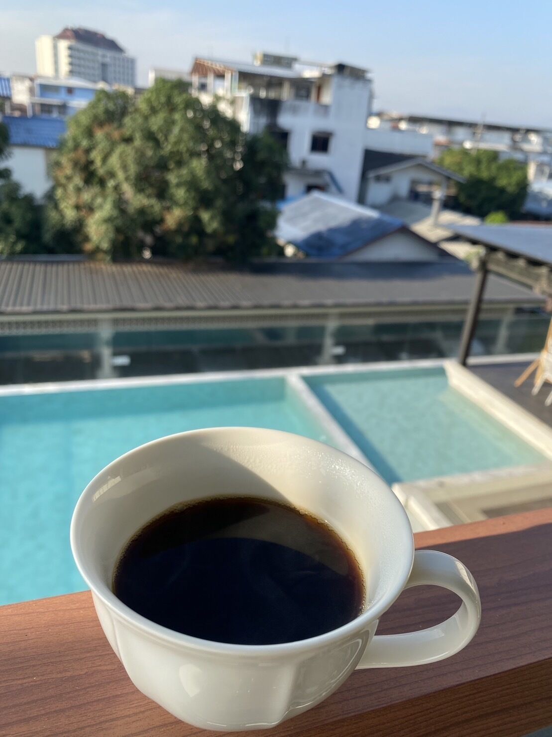 Uma chávena de café branca numa varanda com vista para uma piscina, com edifícios e árvores ao fundo.