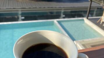 Uma chávena de café branca numa varanda com vista para uma piscina, com edifícios e árvores ao fundo.
