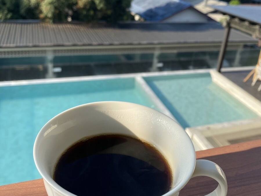 Uma chávena de café branca numa varanda com vista para uma piscina, com edifícios e árvores ao fundo.