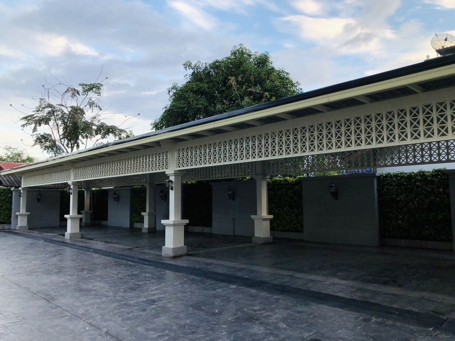 Uma área externa coberta com estrutura de telhado treliçada, colunas e luminárias fixadas na parede.