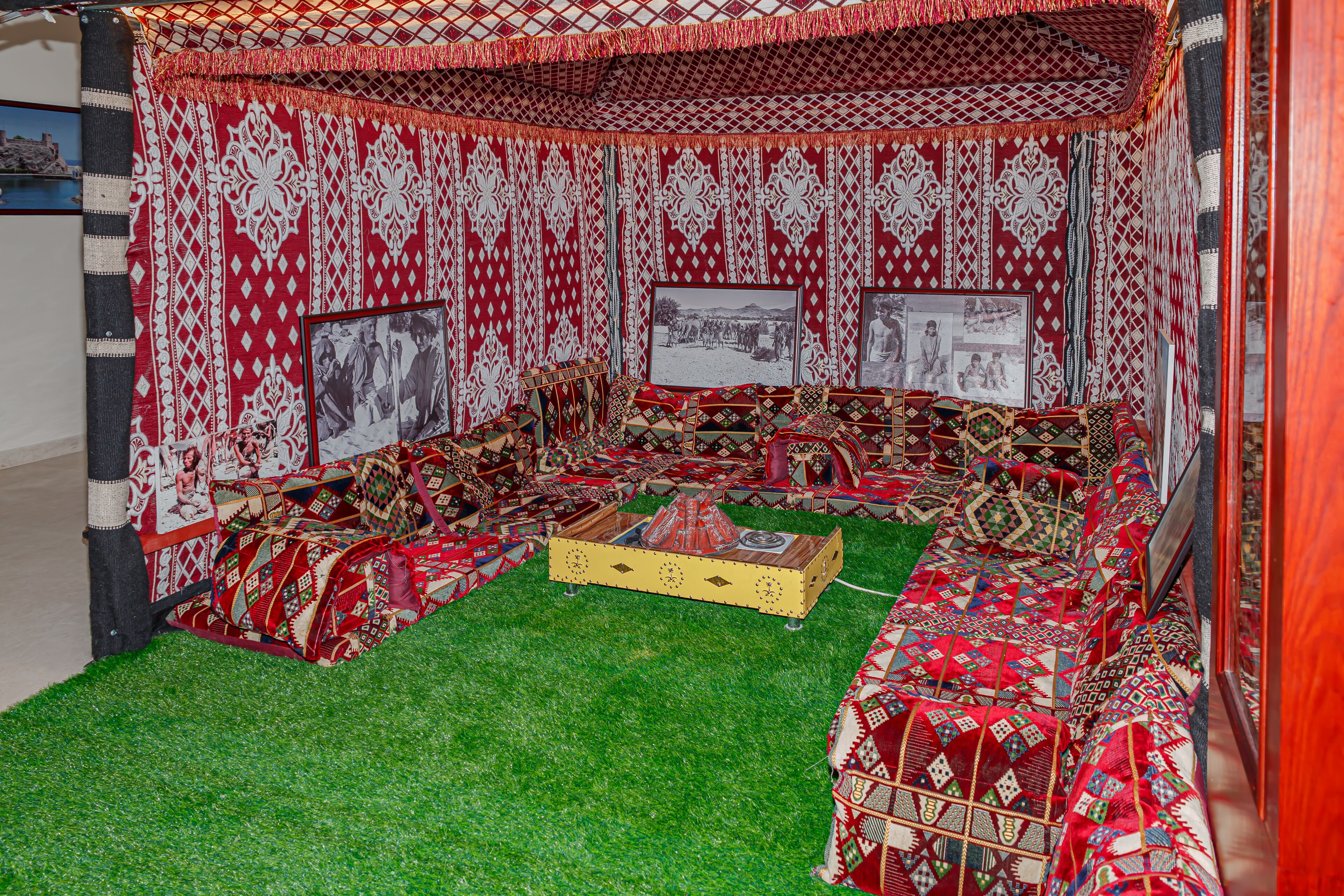 Quarto com carpete vermelho e branco estampado, piso de grama sintética verde e um baú de madeira no centro.