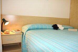 Cama bem arrumada com colcha azul e branca listrada, uma mesinha de cabeceira com abajur e um brinquedo de pelúcia sobre a cama.