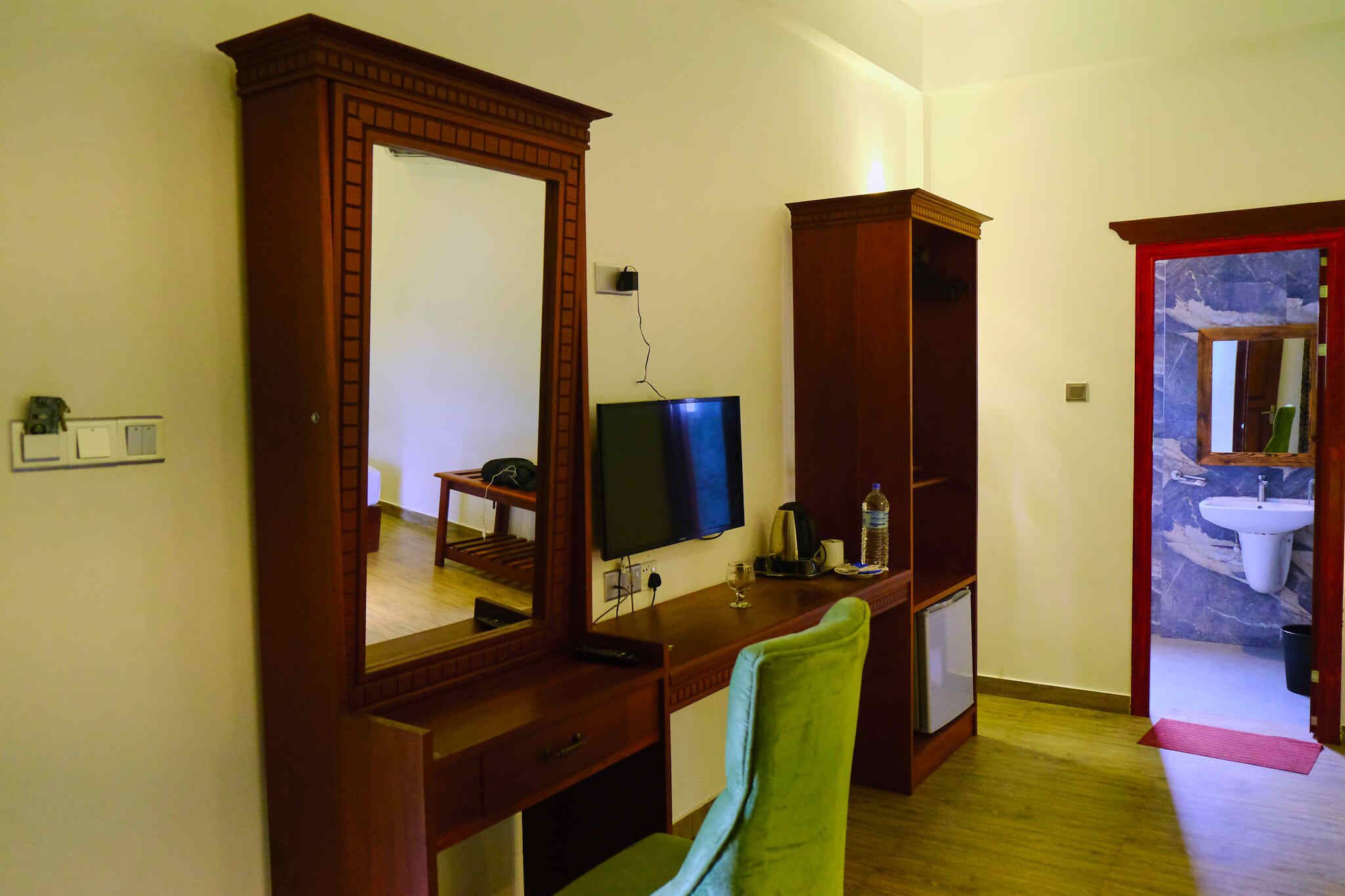 Quarto de hotel com uma escrivaninha de madeira, televisão, espelho e banheiro visível através de uma porta aberta.