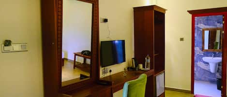 Quarto de hotel com uma escrivaninha de madeira, televisão, espelho e banheiro visível através de uma porta aberta.