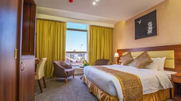Quarto de hotel com cama grande, duas cadeiras, escrivaninha e janela com cortinas.