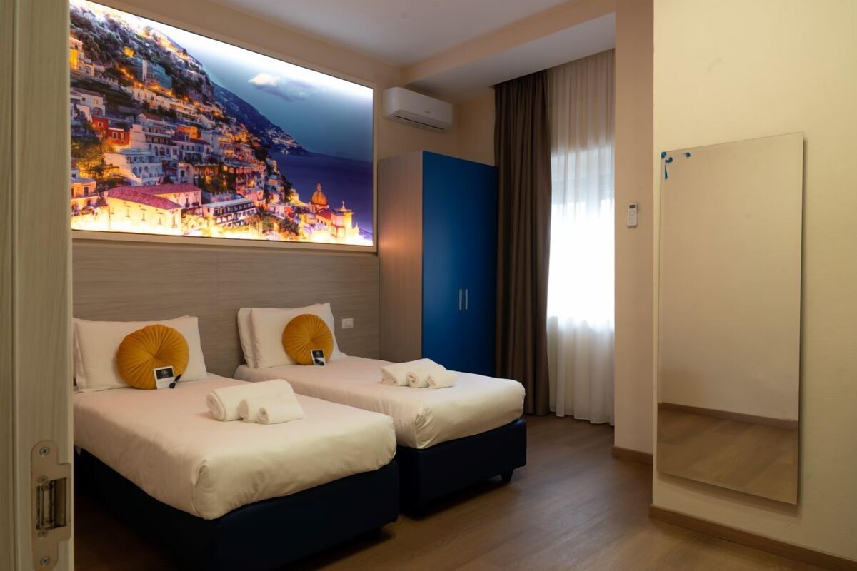 Quarto de hotel com duas camas, uma janela ampla com vista para uma cidade costeira, um guarda-roupa azul e um espelho na parede.