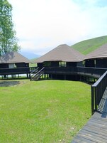 Várias cabanas de madeira com varandas, unidas por um deck de madeira, com colinas e céu claro ao fundo.