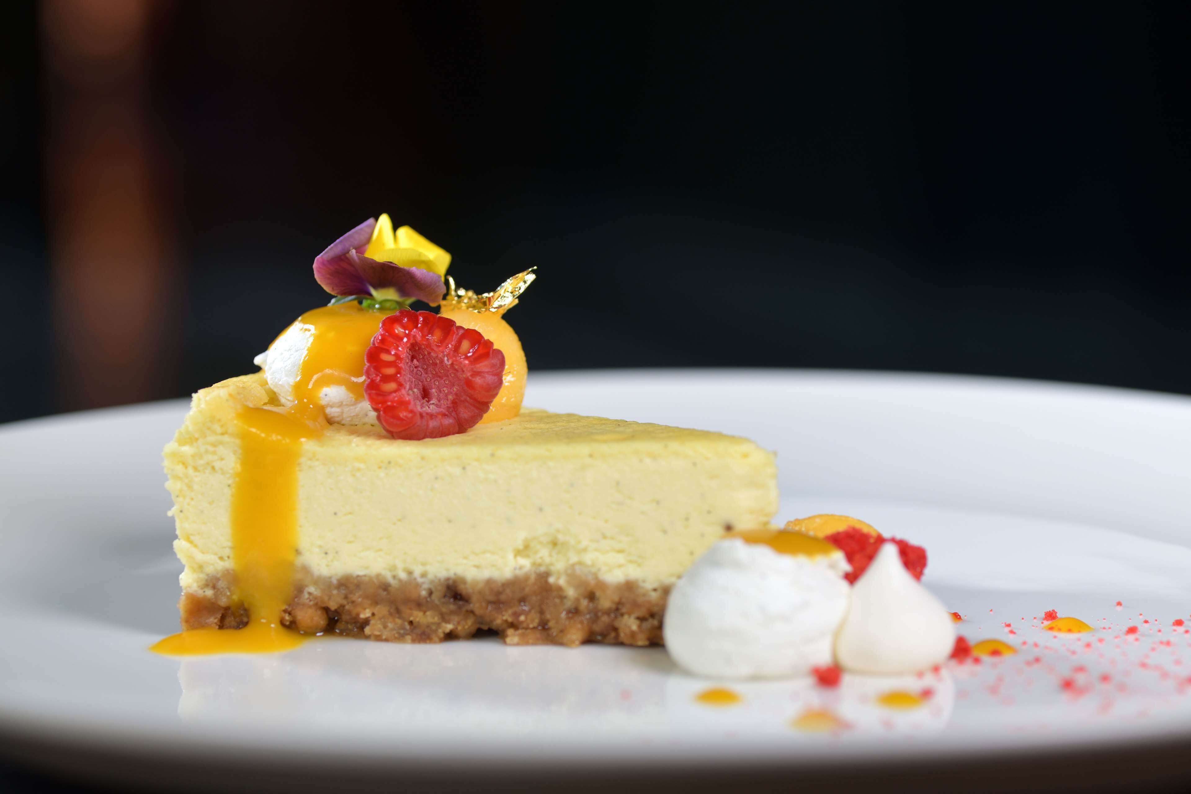 Uma fatia de cheesecake com um fio de caramelo dourado, coberta com uma morango, um pouco de creme e uma folha decorativa.