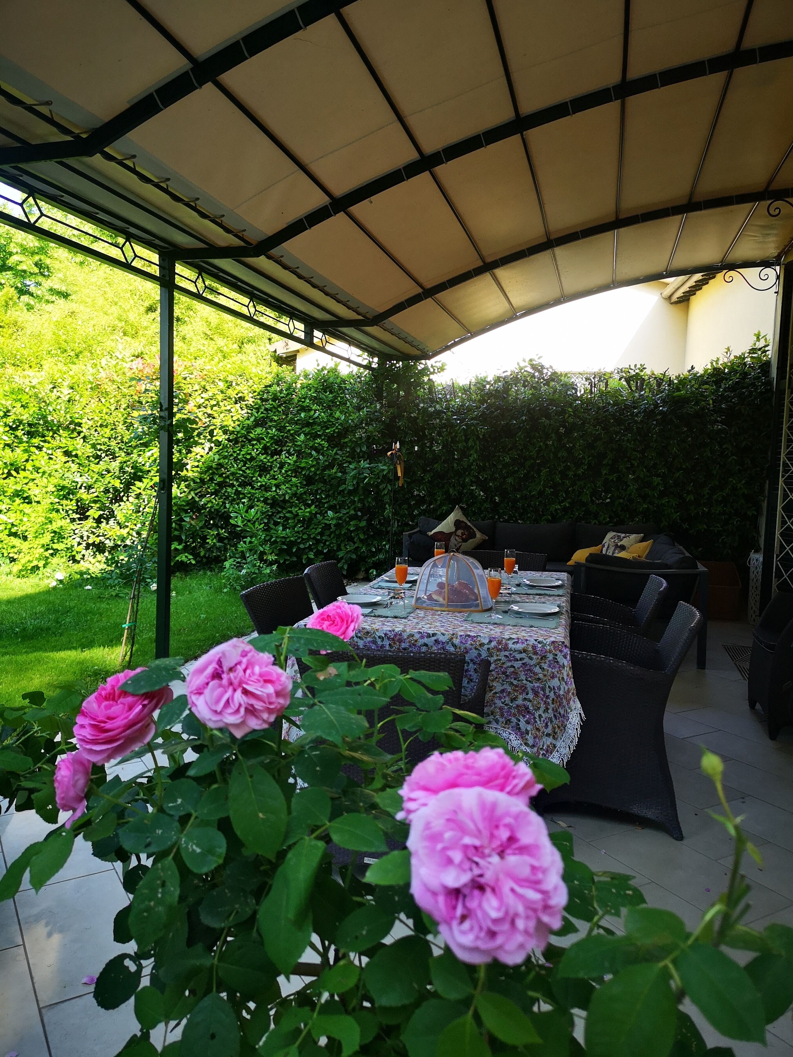 Um pátio externo com uma mesa posta para dois, cercado por vegetação exuberante e flores cor-de-rosa.
