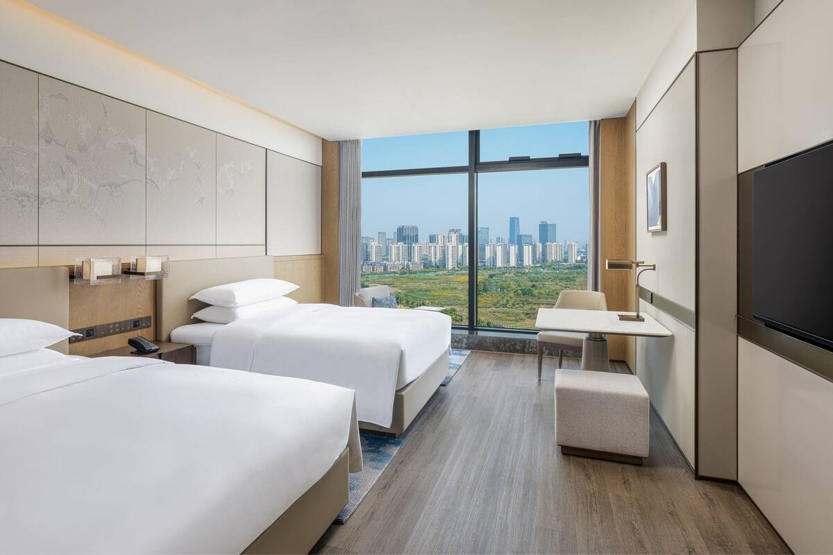 Quarto de hotel com duas camas, uma mesa de trabalho e uma grande janela com vista para a cidade.