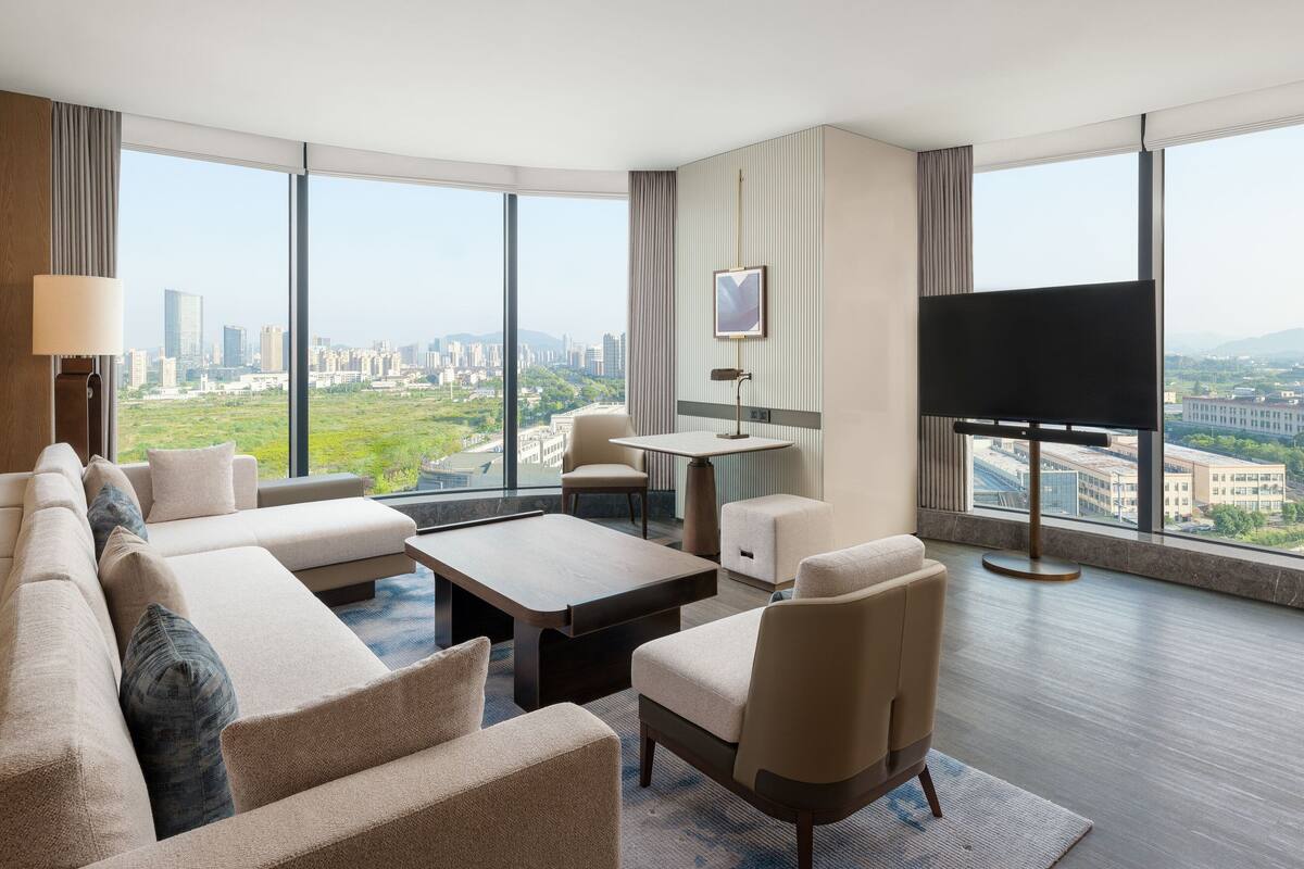 Quarto de hotel moderno com um sofá grande, uma mesa de centro, TV e vista para a cidade.