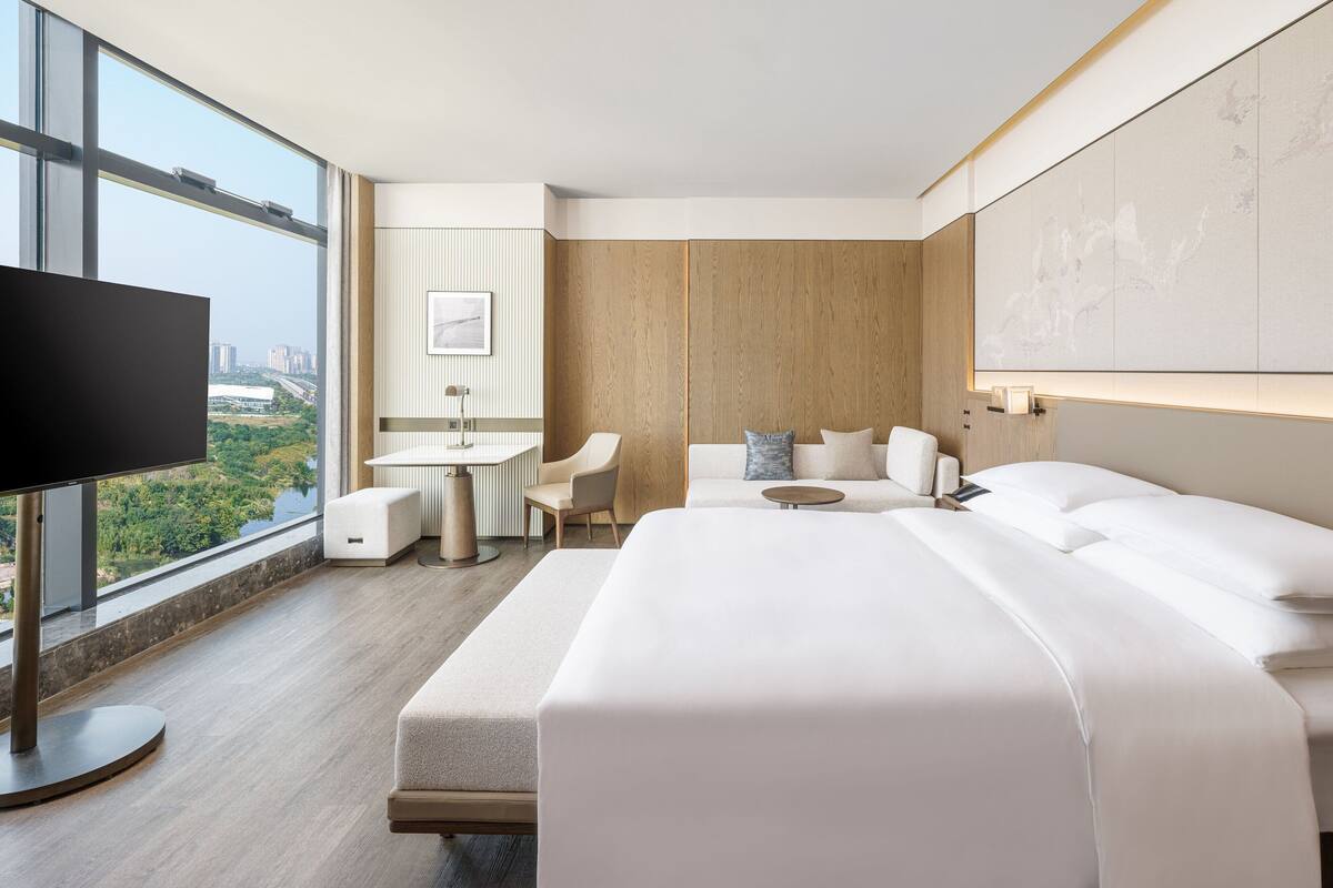 Quarto de hotel moderno com uma cama grande, uma mesa e uma cadeira. Há uma televisão no lado esquerdo do quarto.