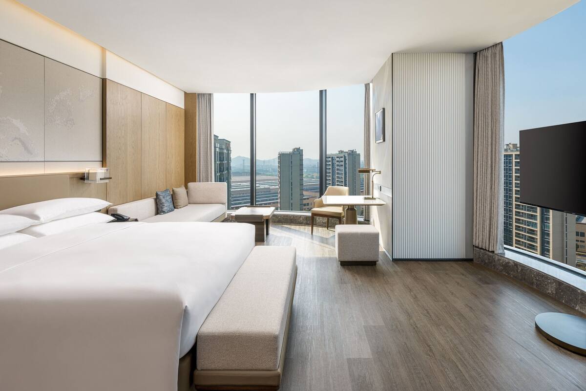 Quarto de hotel moderno com uma cama grande, um sofá, uma mesa pequena e uma televisão. O quarto tem janelas grandes com vista para a cidade.