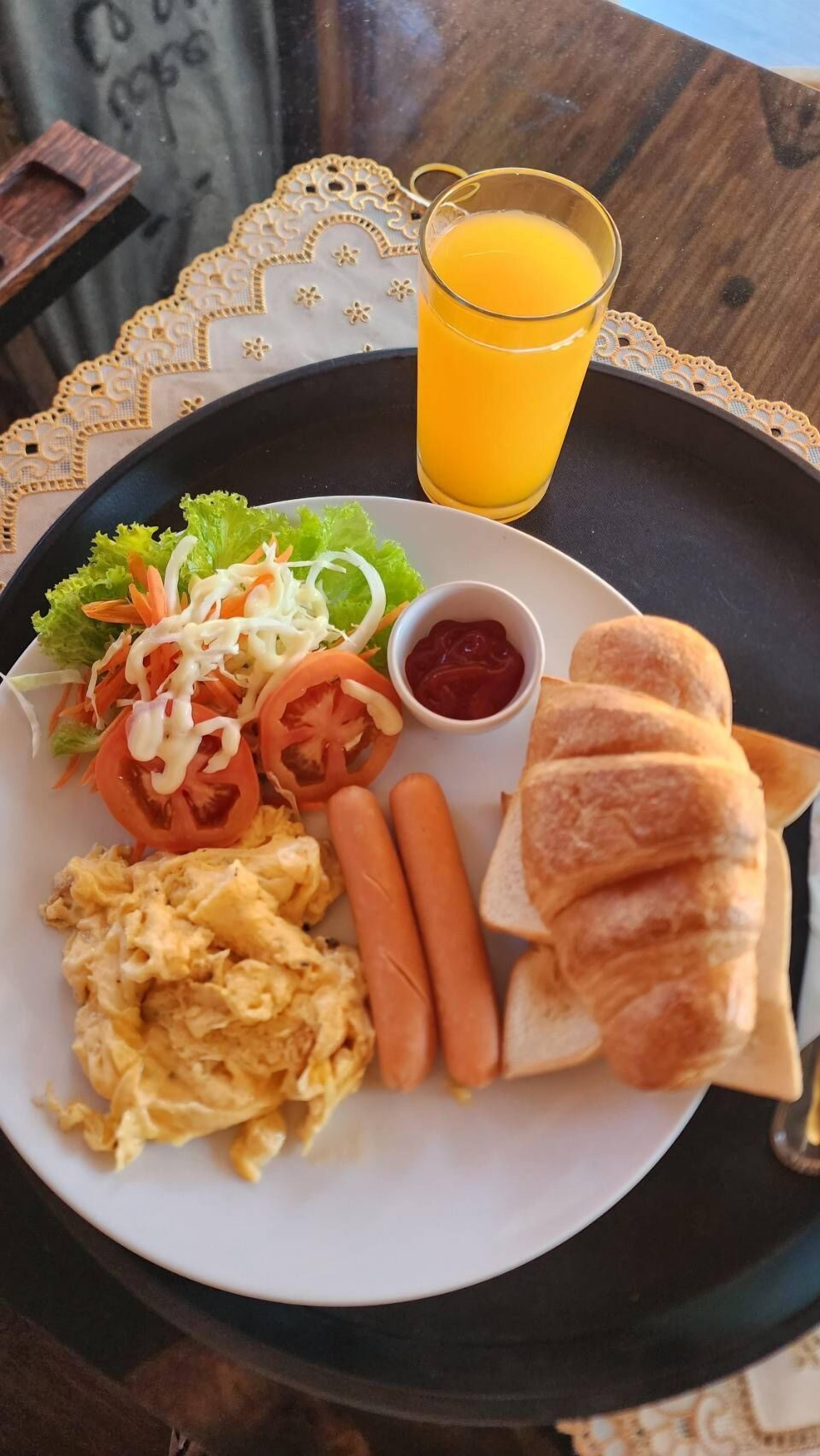 Uma bandeja com um prato de comida, incluindo ovos mexidos, linguiça, salada e um croissant, além de um copo de suco de laranja.