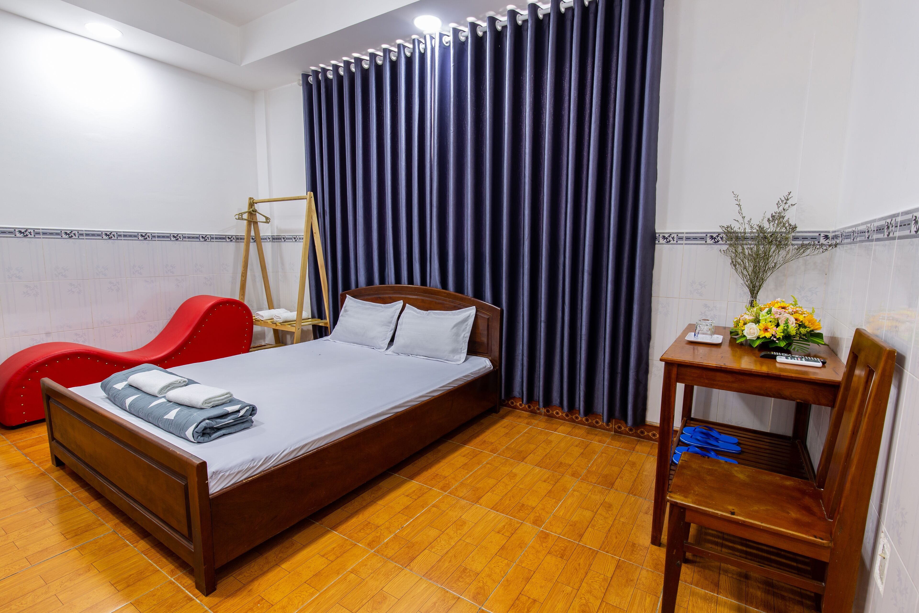 Cama bem arrumada com almofadas e toalha dobrada, uma cadeira de madeira, uma pequena mesa com um vaso de flores e uma cadeira vermelha.