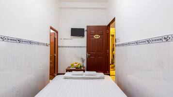 Quarto de hotel com uma cama, porta de madeira com o número 204, uma mesinha de centro com um vaso de flores e uma televisão fixada na parede.