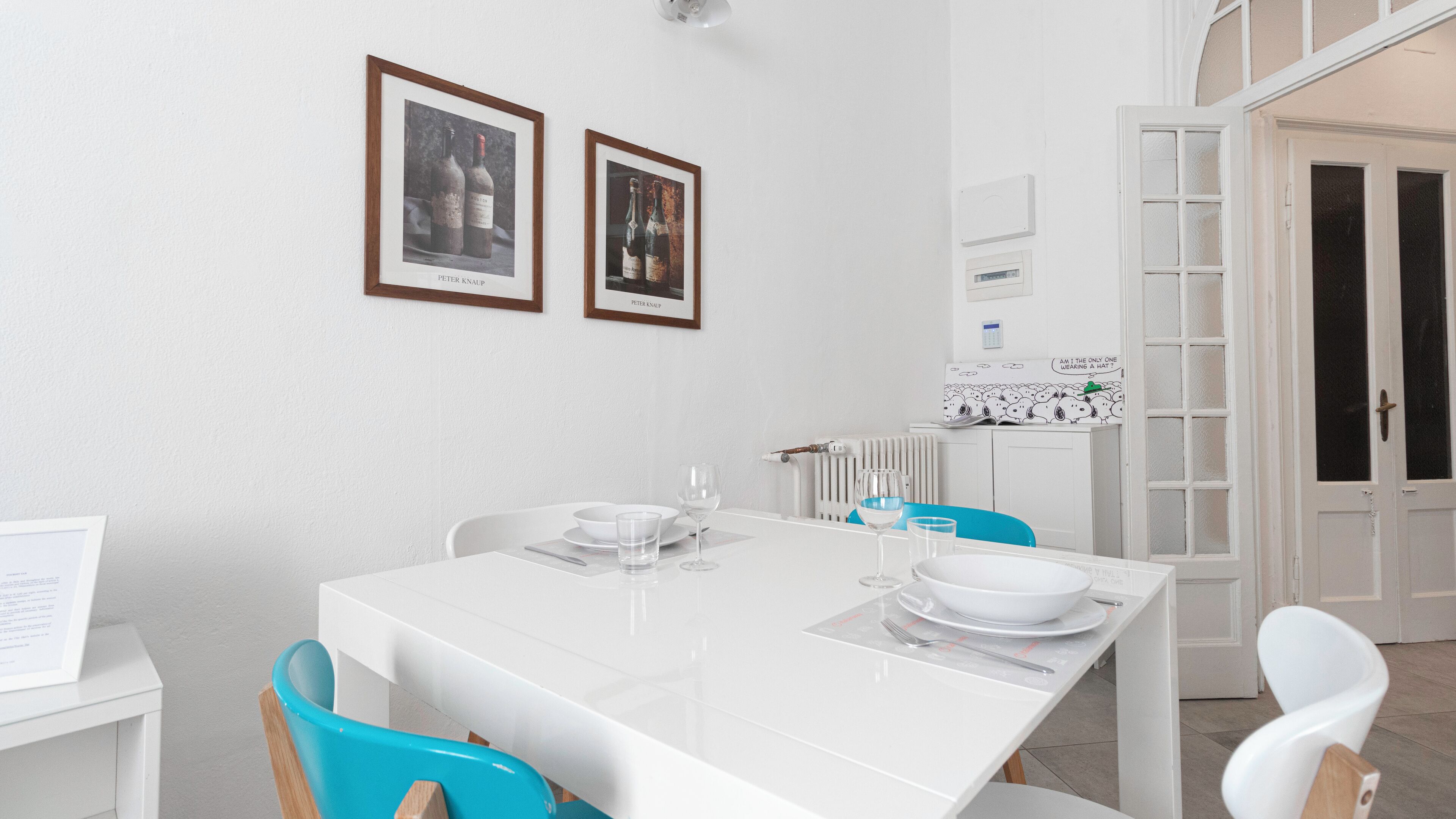 Uma área de refeições com mesa branca posta para dois, cadeiras azuis e quadros nas paredes.
