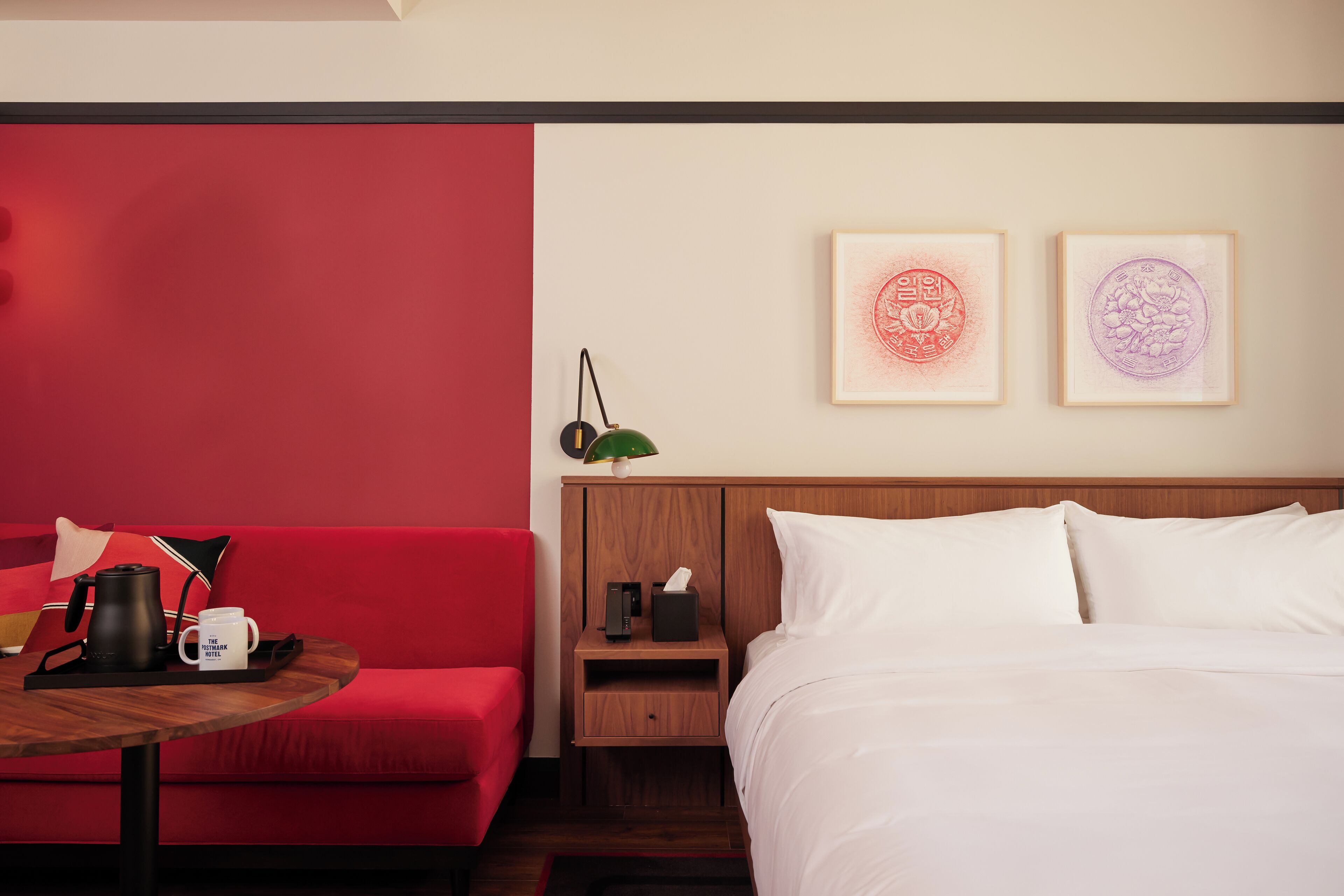 Quarto de hotel com um sofá vermelho, uma mesa de cabeceira de madeira com uma luminária, uma cama com roupa de cama branca e duas obras de arte emolduradas na parede.