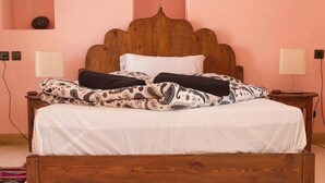 Uma cama de madeira com lençol branco e manta estampada, duas luminárias de cabeceira e uma parede rosa.