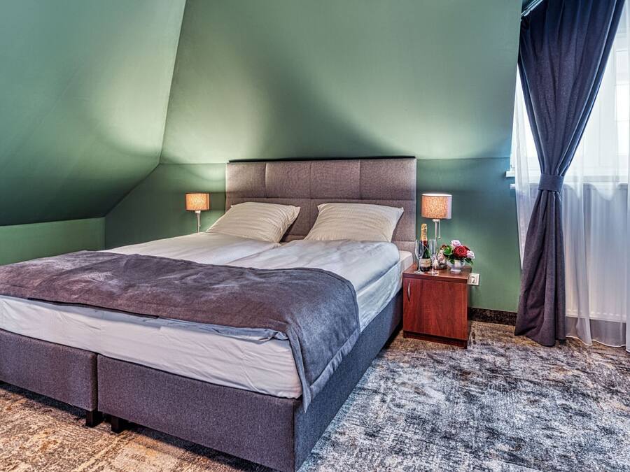 Quarto com cama grande, mesa de cabeceira, abajur, uma garrafa de champanhe e um vaso de flores.