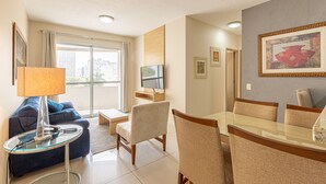 Uma sala de estar moderna com um sofá, uma mesa de centro de vidro, uma mesa de jantar com cadeiras, uma TV e uma janela grande com cortinas.