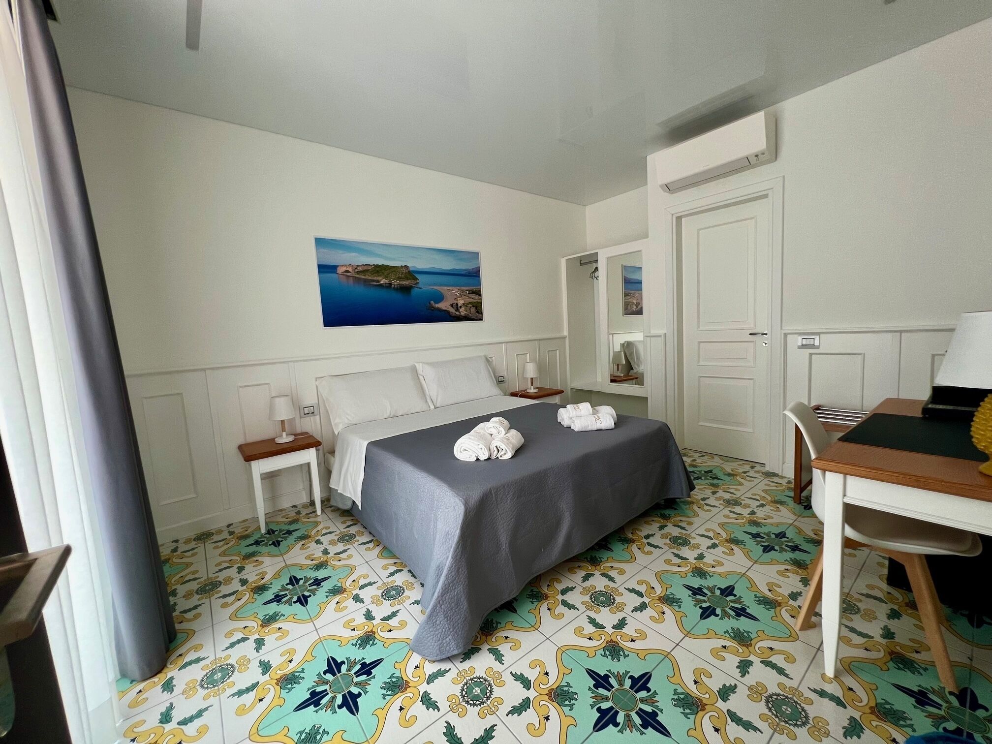 Quarto de hotel com cama, mesas de cabeceira, escrivaninha e uma cadeira.