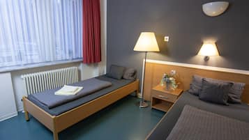 Quarto de hotel com uma cama de solteiro, uma cama menor, uma mesinha de cabeceira com luminária, um criado-mudo, uma janela com cortinas e um ponto de luz fixado na parede.