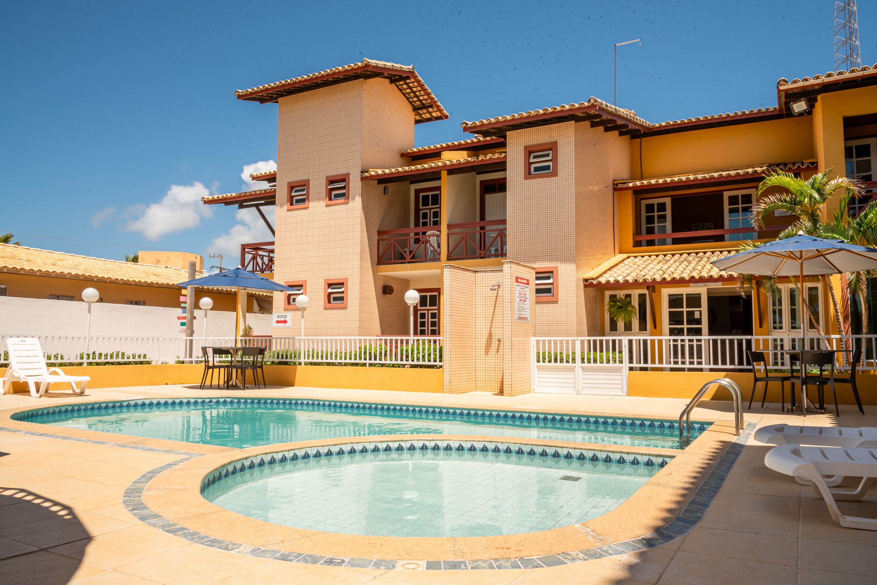 Piscina com espreguiçadeiras, prédio com varandas e um céu azul límpido.