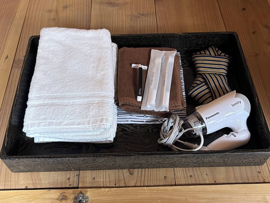 Uma caixa de vime preta contém uma toalha branca, uma toalha marrom, um travesseiro listrado, um secador de cabelo Panasonic e um conjunto de acessórios para modelar o cabelo.