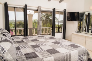 Quarto com colcha xadrez, varanda com cortinas, televisão na parede e vista para o verde lá fora.