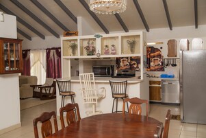 Uma área de refeições com mesa e cadeiras de madeira, uma cozinha com geladeira, micro-ondas e armários, e uma sala de estar com sofá e cortinas.