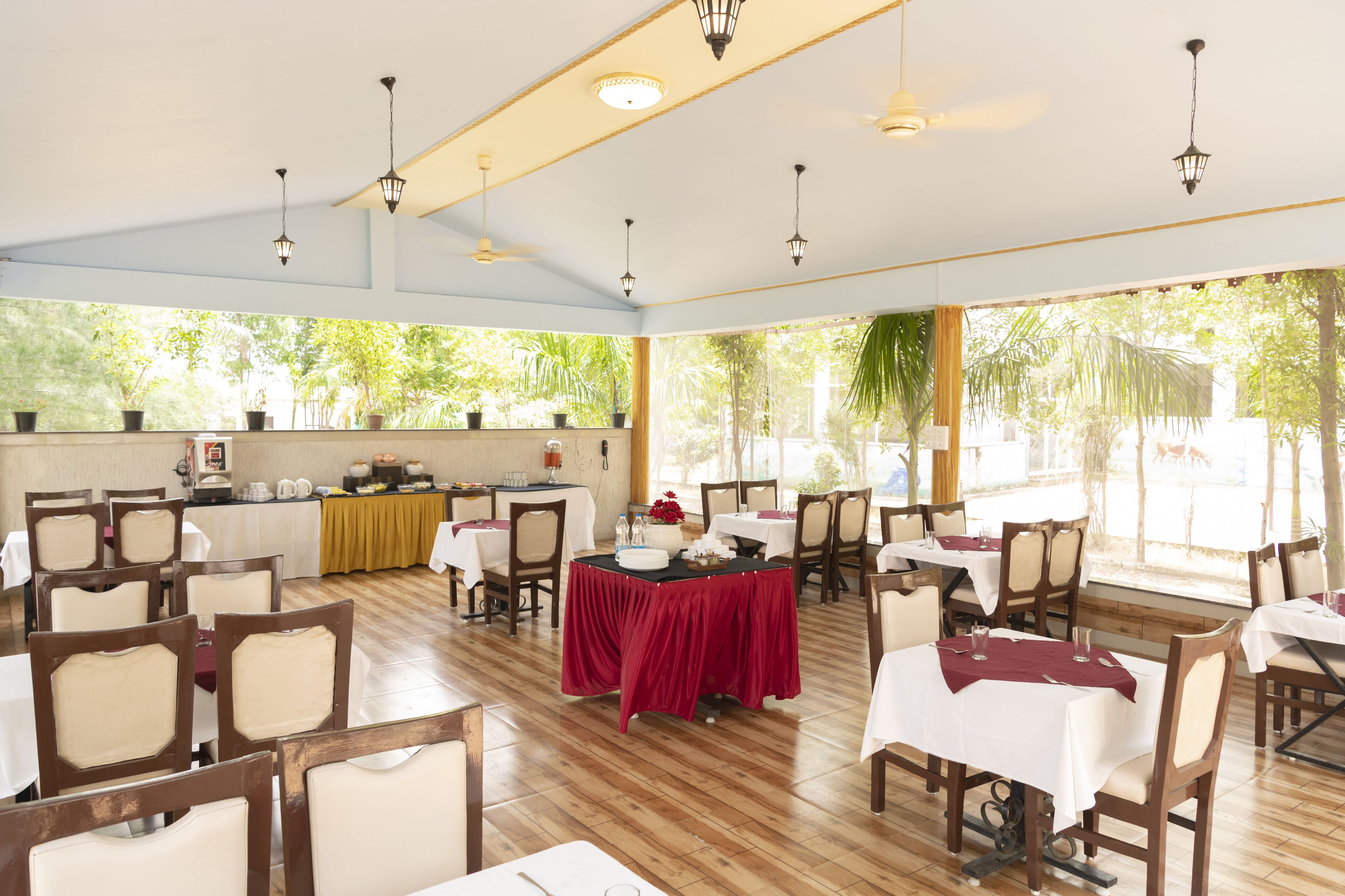 Uma ampla sala de jantar com várias mesas e cadeiras, janelas grandes com vista para o verde e teto com luminárias pendentes.