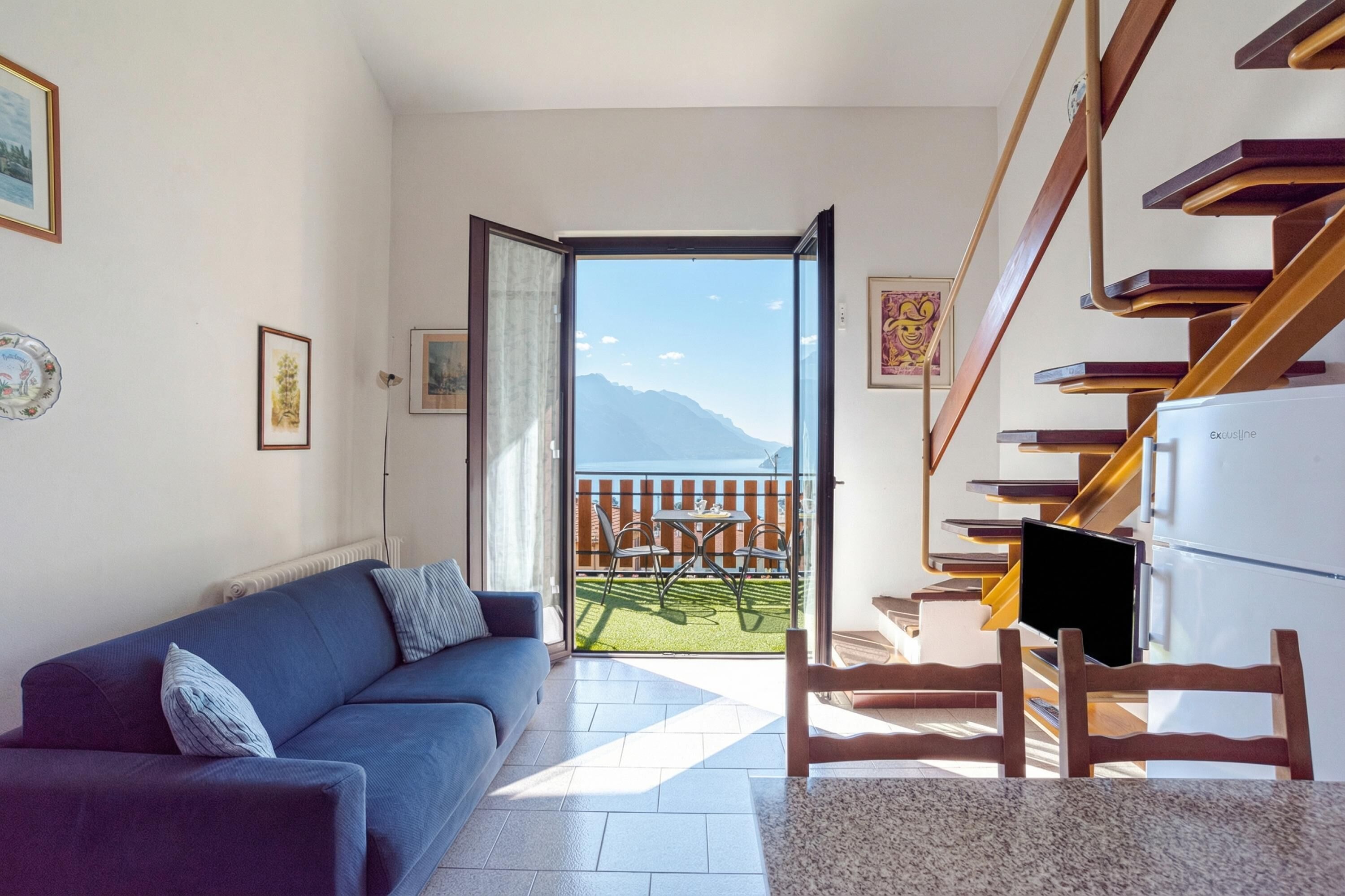 Um quarto com um sofá azul, uma mesa de jantar com cadeiras, uma escada e vista para montanhas por uma porta aberta.