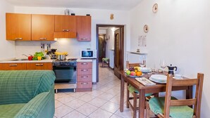Cozinha compacta com armários de madeira, fogão e micro-ondas. Possui um espaço de jantar com mesa posta para dois e um sofá verde.