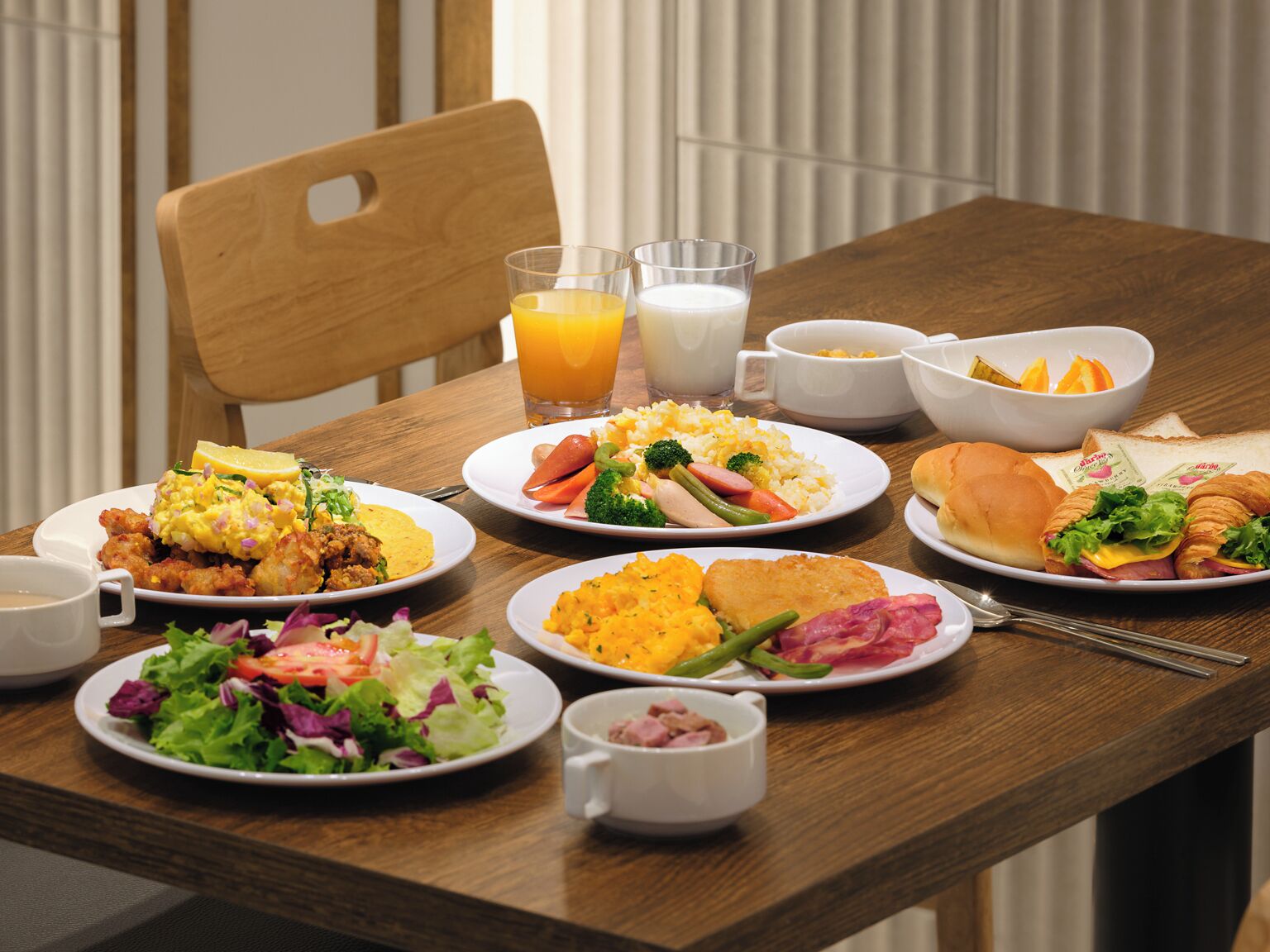 Uma mesa de jantar com diversos pratos, incluindo ovos mexidos, uma salada e sanduíches. Há também copos de suco de laranja e leite.