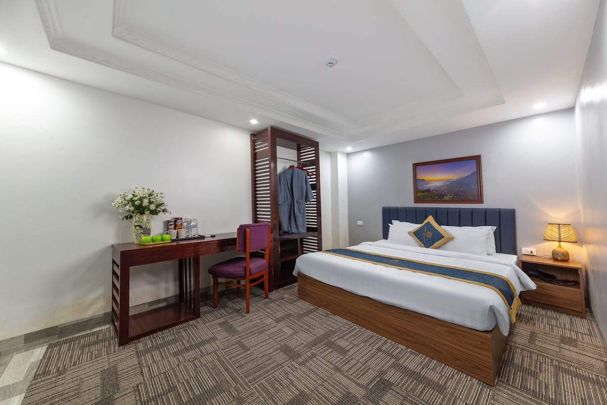 Quarto de hotel com cama, escrivaninha, cadeira, mesa de cabeceira e prateleira na parede.