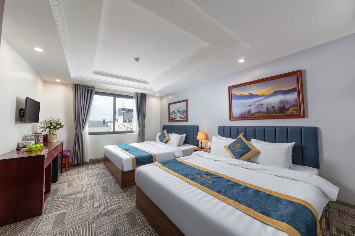 Quarto de hotel com duas camas, uma escrivaninha, uma televisão e uma janela com cortinas.