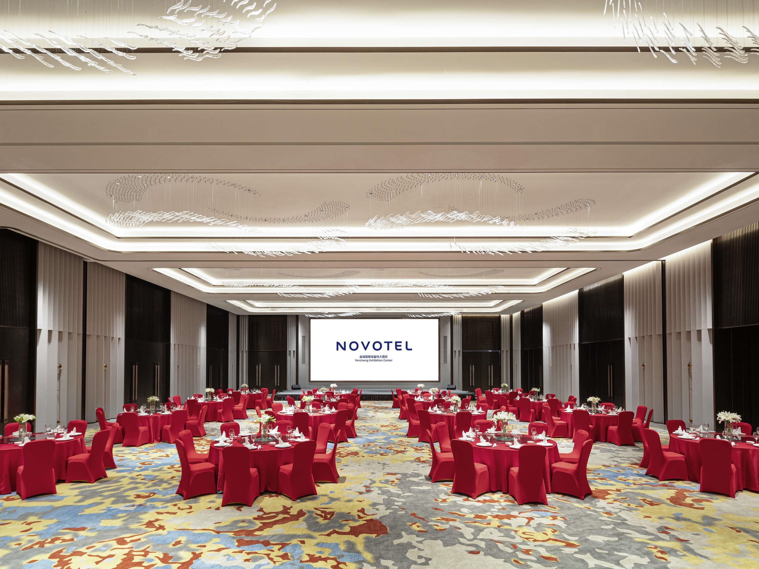 Uma sala de conferências com cadeiras e mesas vermelhas, uma tela grande exibindo 'NOVOTEL' e um carpete estampado.