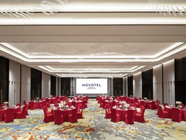 Uma sala de conferências com cadeiras e mesas vermelhas, uma tela grande exibindo 'NOVOTEL' e um carpete estampado.