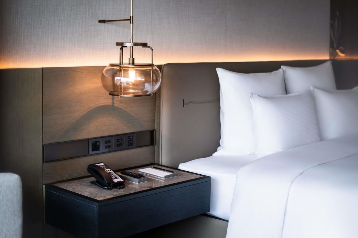 Quarto de hotel moderno com uma cama, mesa de cabeceira com telefone e controles remotos, e luminária pendente única.