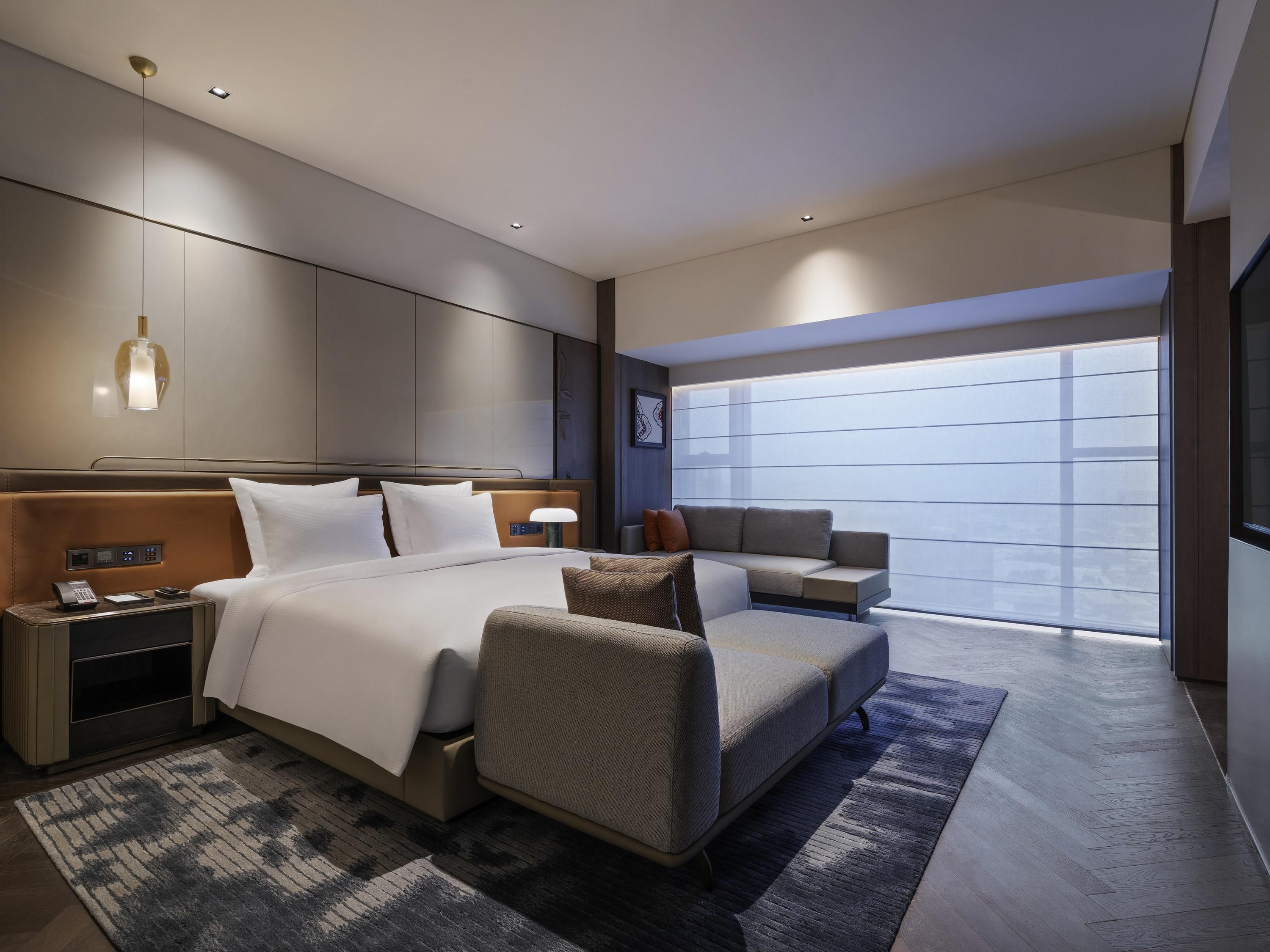 Quarto de hotel moderno com uma cama grande, um sofá, uma escrivaninha e uma janela ampla.