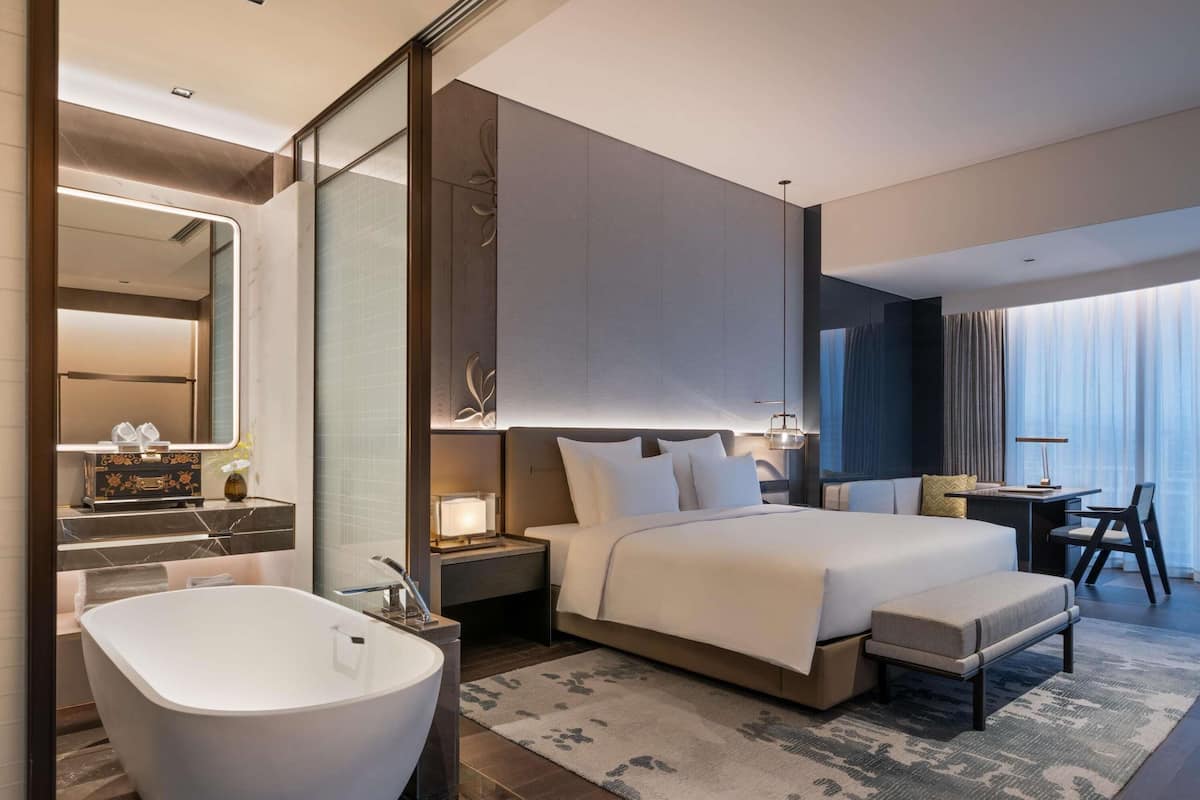 Quarto de hotel moderno com uma cama grande, uma área de estar separada, uma banheira e um espelho amplo.