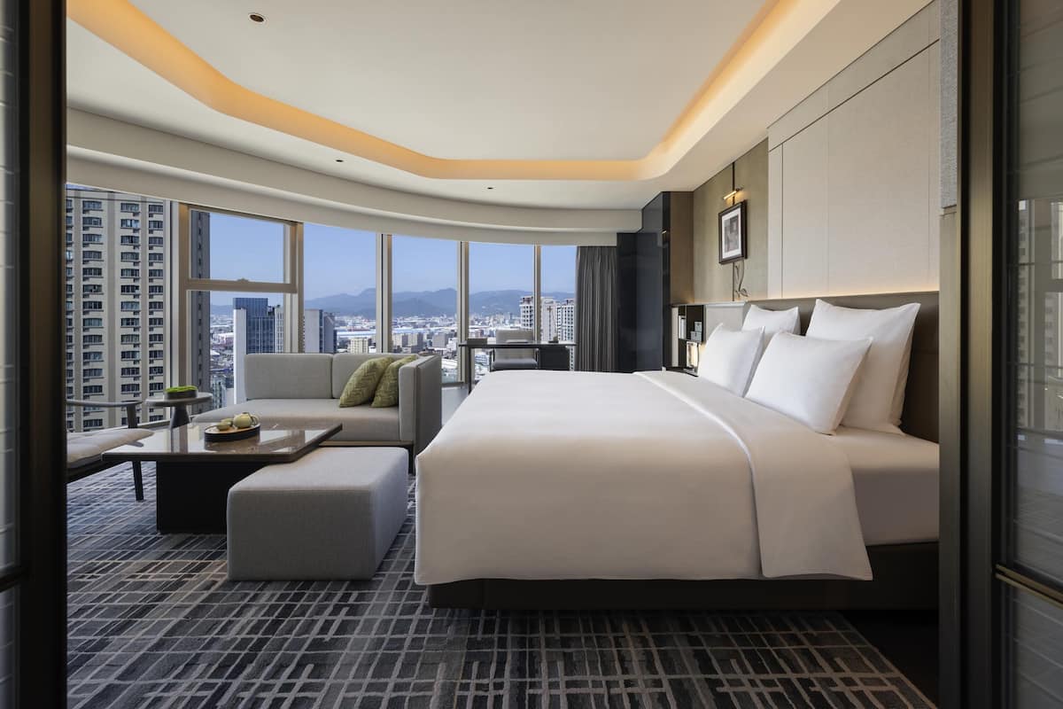 Quarto de hotel com uma cama grande, um sofá, uma mesa pequena com bandeja e vista para a cidade através de janelas amplas.