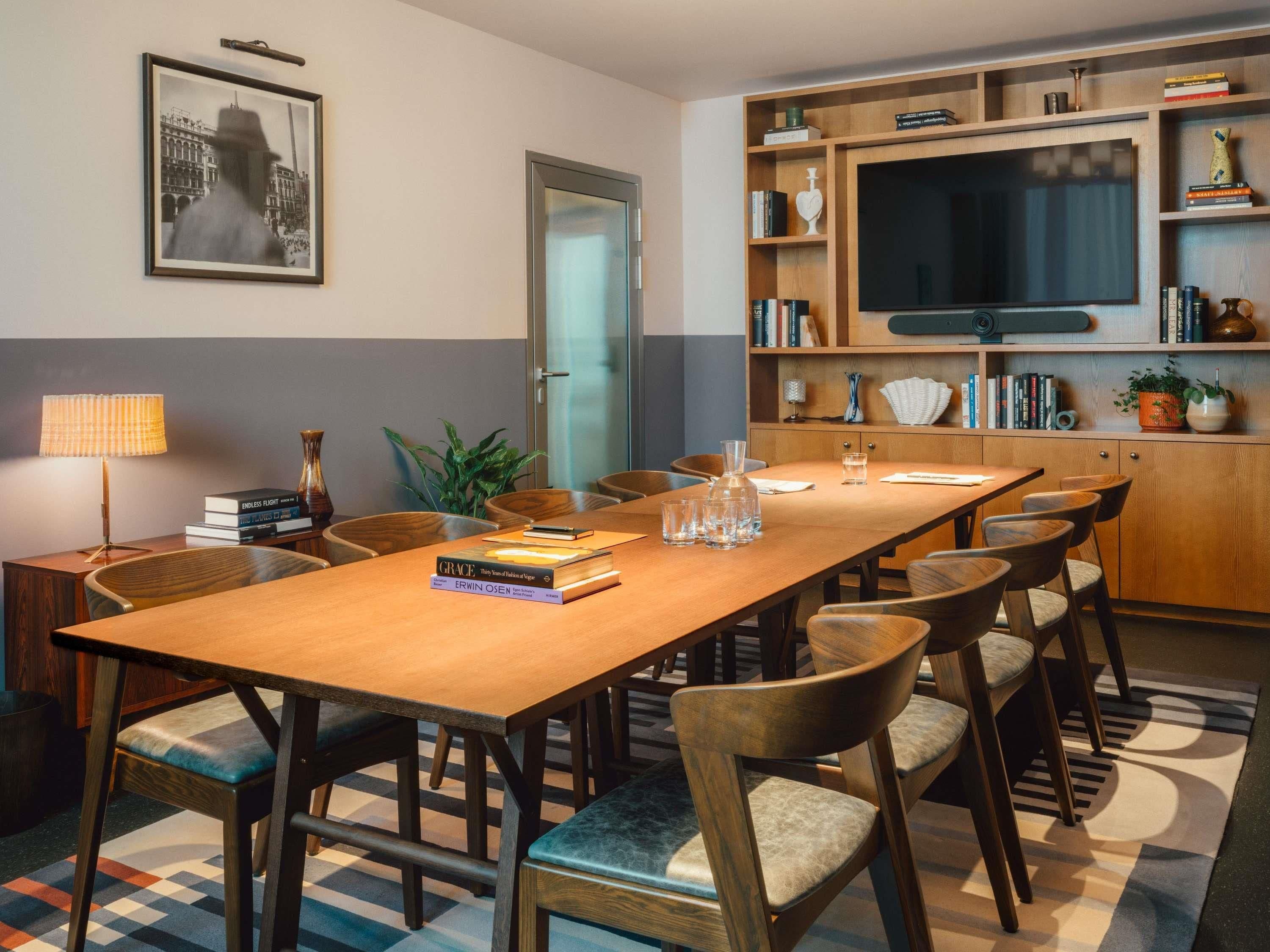 Uma sala de conferência com uma mesa comprida, cadeiras, uma estante de livros, uma televisão e um quadro na parede.