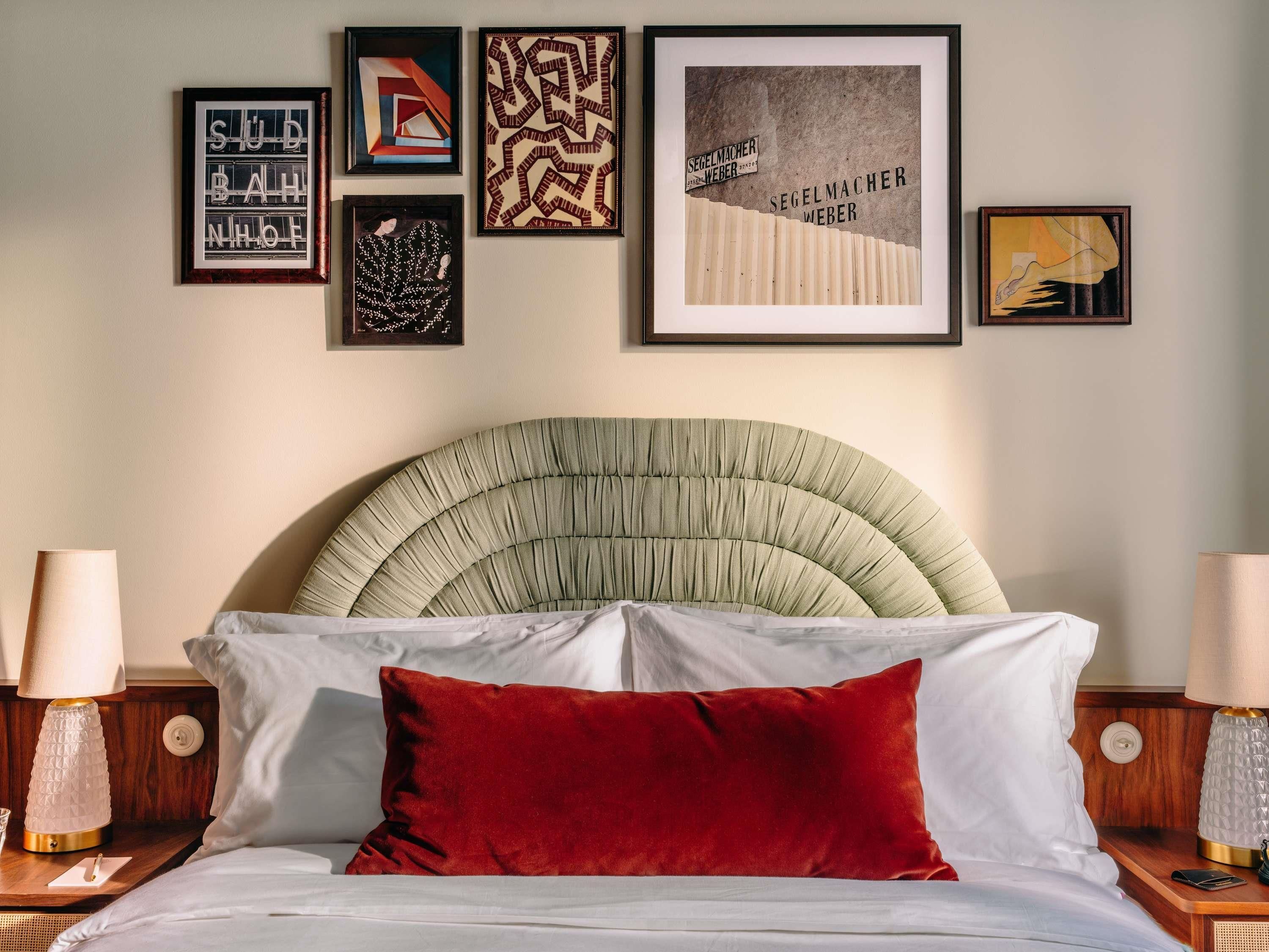 Cama bem arrumada com cabeceira estofada verde, roupa de cama branca e travesseiro vermelho. Há quadros na parede acima da cama.
