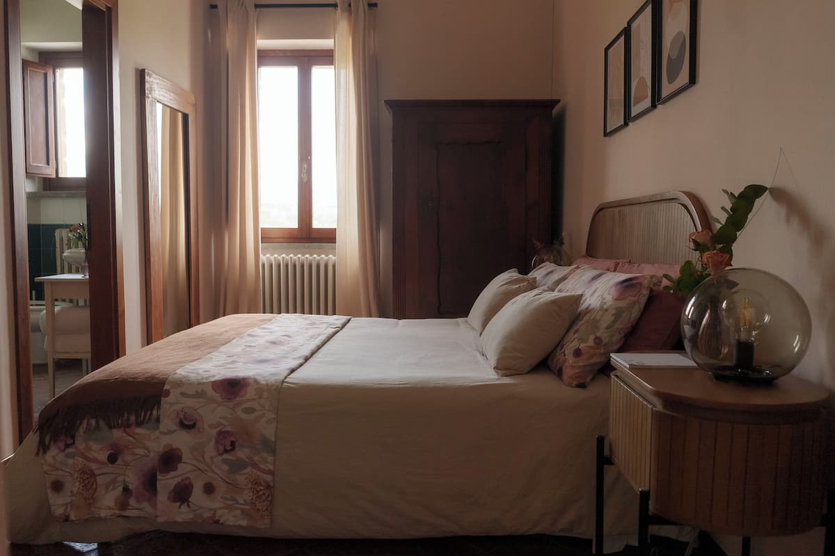 Quarto com cama, mesa de cabeceira com abajur, janela com cortinas e radiador.