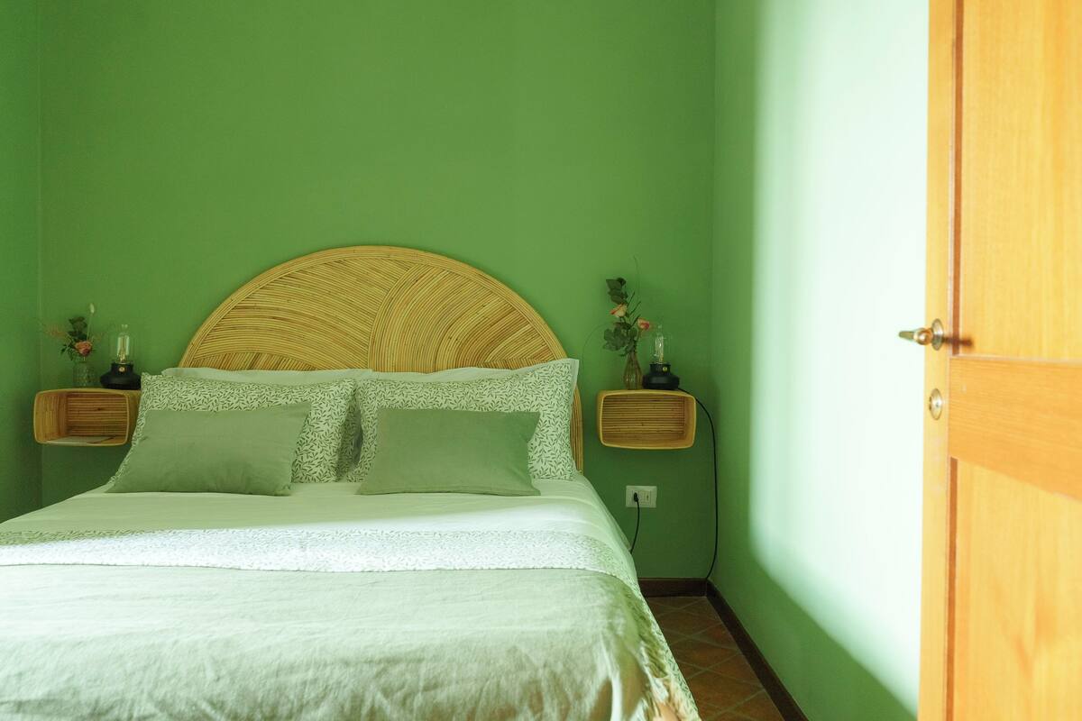 Uma cama com cabeceira de tecido, duas mesas de cabeceira com abajures e uma parede verde.