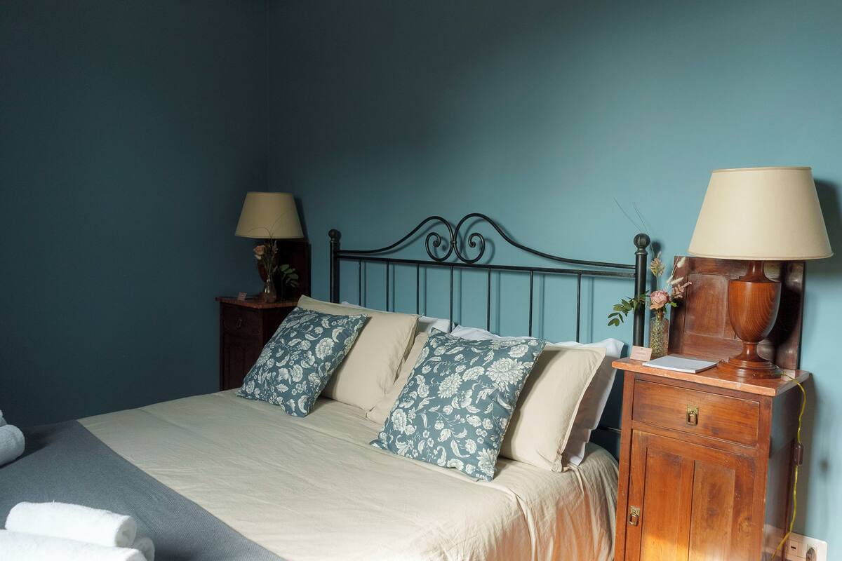 Cama bem arrumada com almofadas florais, mesa de cabeceira de madeira com abajur e parede azul.